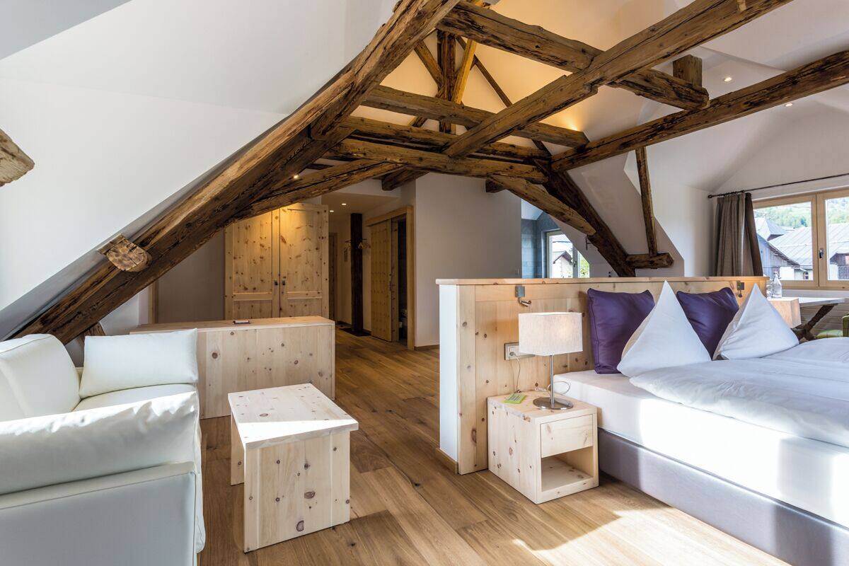 Quarto estilo loft com vigas de madeira aparente, cama branca e mesa de cabeceira de madeira.
