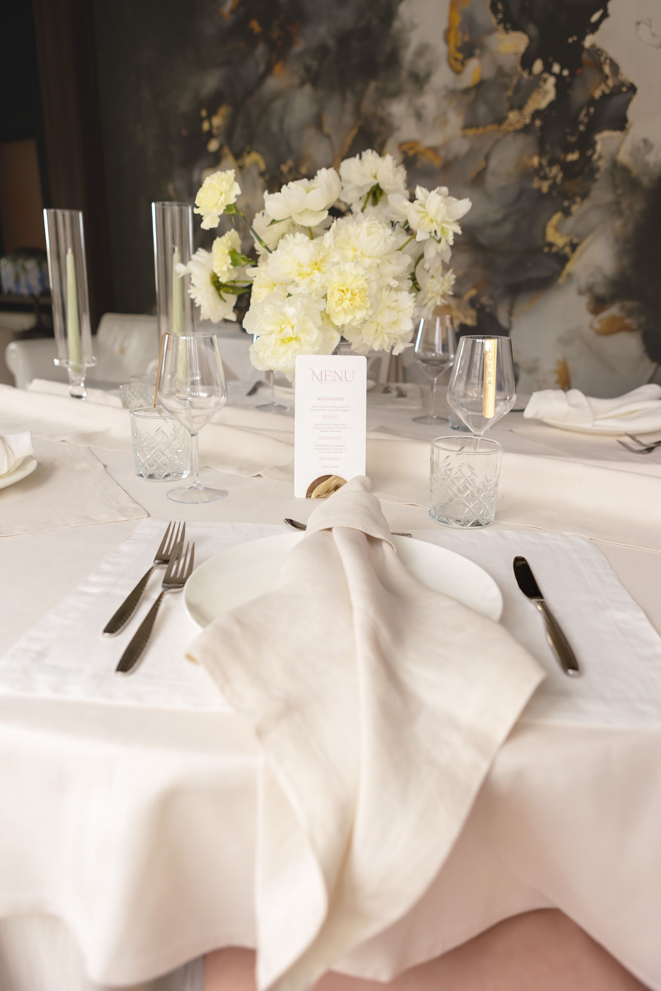 Mesa posta para refeição, com menu, copos de vidro transparente e um vaso de flores brancas.
