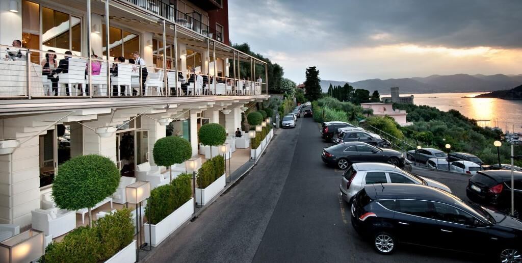 Um hotel com área de estar coberta ao ar livre, uma fileira de carros estacionados e vista para a água e montanhas ao fundo.