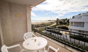 Varanda com mesa redonda e cadeiras, com vista para uma praia e uma estrada com vegetação e edifícios.