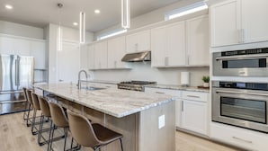 Cozinha moderna com armários brancos, uma ilha grande com banquetas, eletrodomésticos de aço inoxidável e iluminação pendente.
