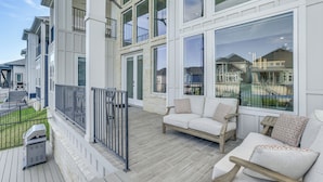 Varanda ampla com deck de madeira, mobiliário de exterior branco e grandes janelas com vista para as casas vizinhas.