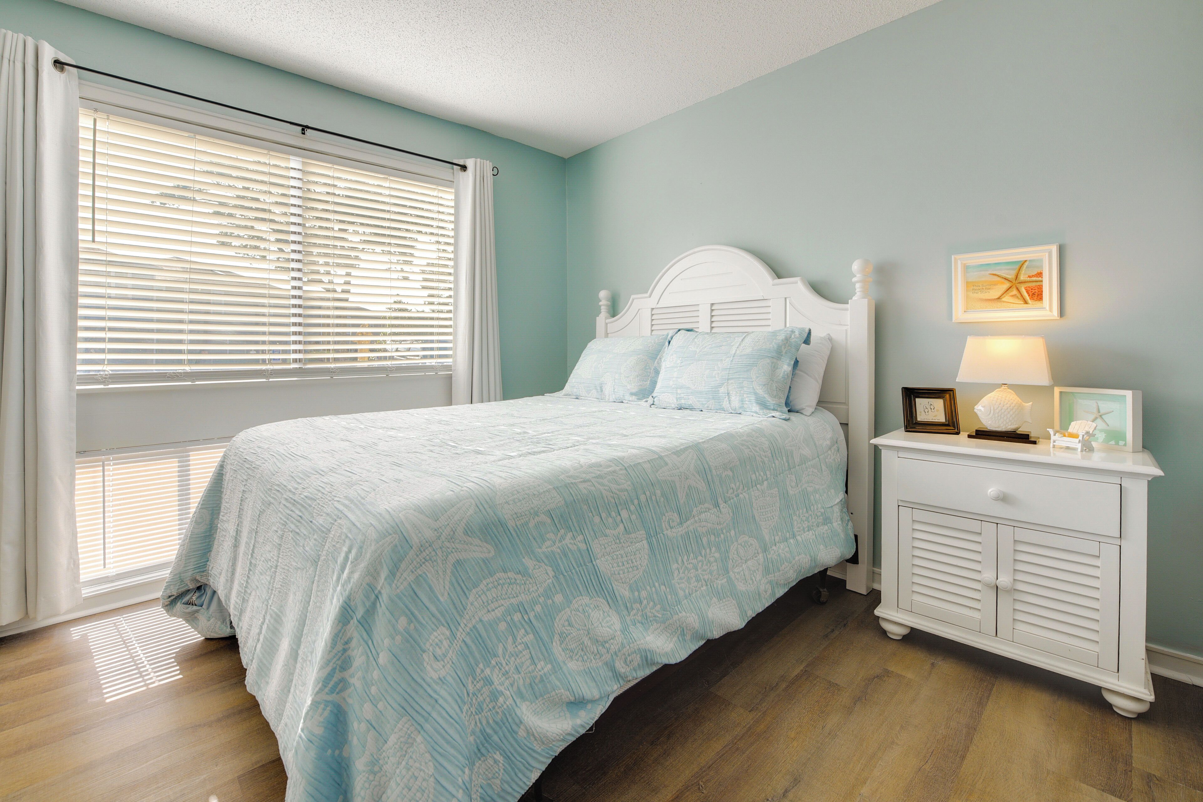 Quarto com cama, mesa de cabeceira, janela com persianas e um quadro pendurado na parede.