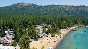 Uma área costeira com praia, carros estacionados e uma colina arborizada ao fundo.