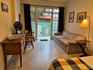 Quarto com sofá, escrivaninha com ventoinha, cadeira, luminária e vista através de uma porta de vidro.