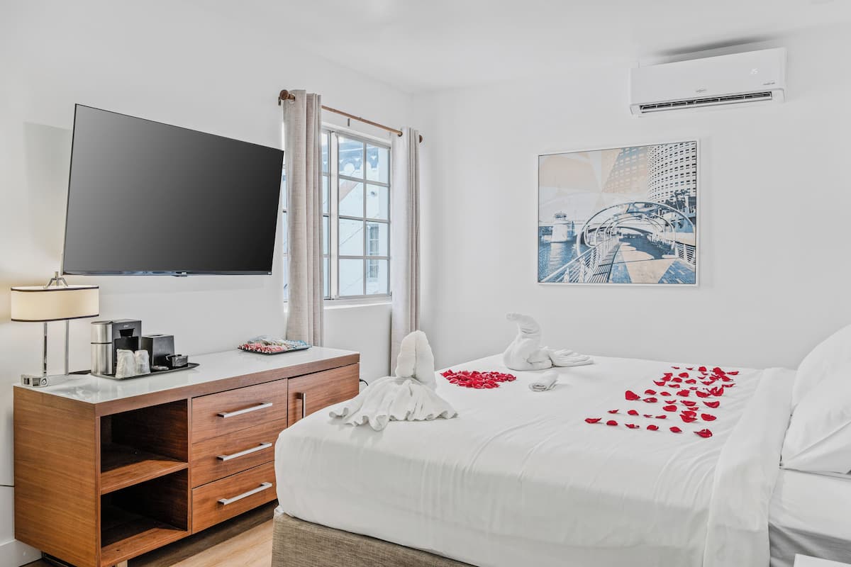 Quarto de hotel com televisão de tela plana, cômoda de madeira, cama com roupa de cama branca e pétalas de rosa, um quadro e um aparelho de ar condicionado.