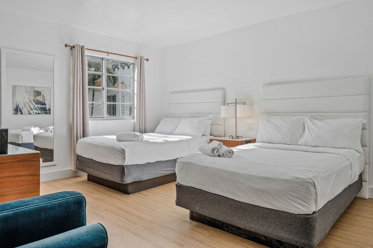 Quarto de hotel com duas camas, uma escrivaninha, uma cadeira e uma janela com cortinas.