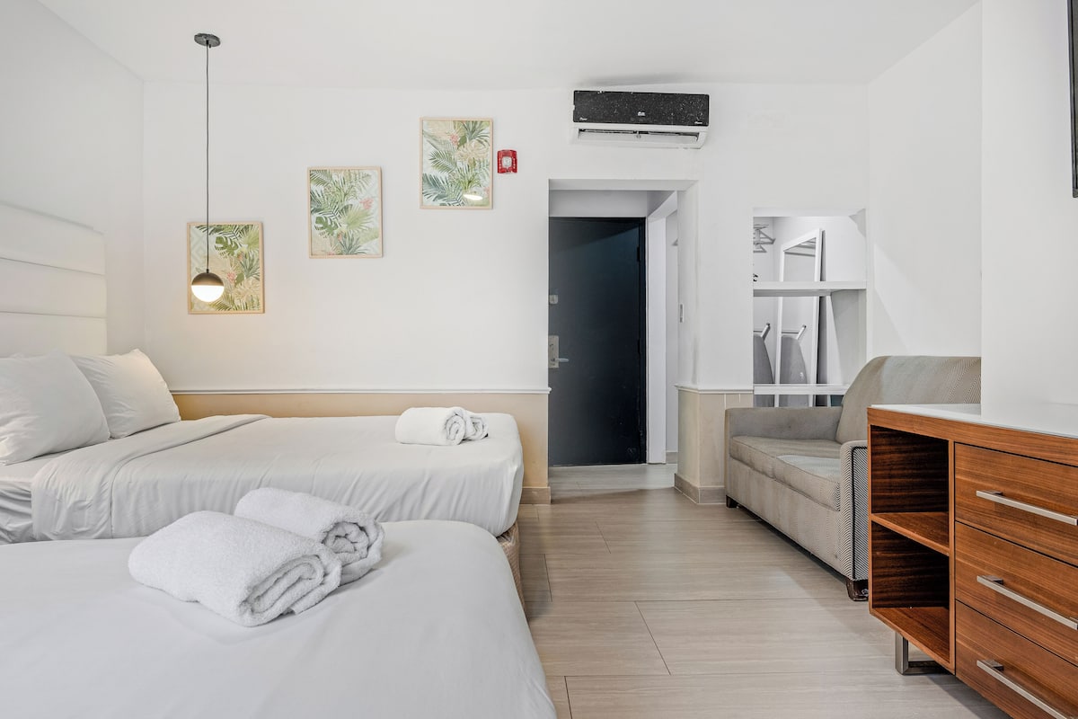 Quarto de hotel com duas camas, cômoda de madeira, sofá e porta que dá para outro espaço.