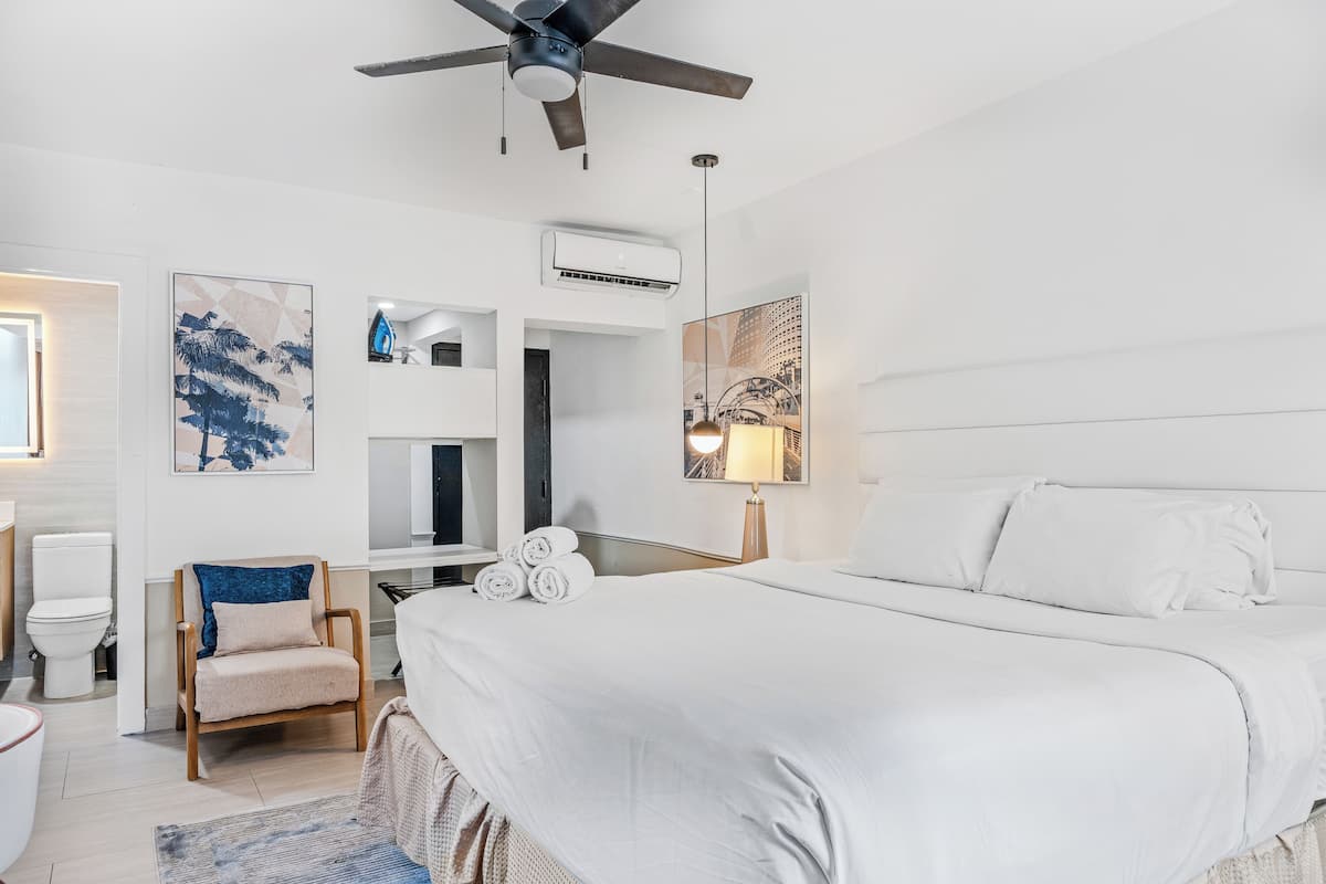Quarto de hotel com uma cama, uma cadeira, uma escrivaninha, ar condicionado e ventoinha no teto.
