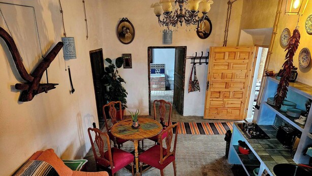 Uma sala de estar aconchegante com uma mesa redonda de madeira, cadeiras vermelhas, um sofá com almofadas estampadas, um lustre e uma área de cozinha com armários azuis e um fogão.