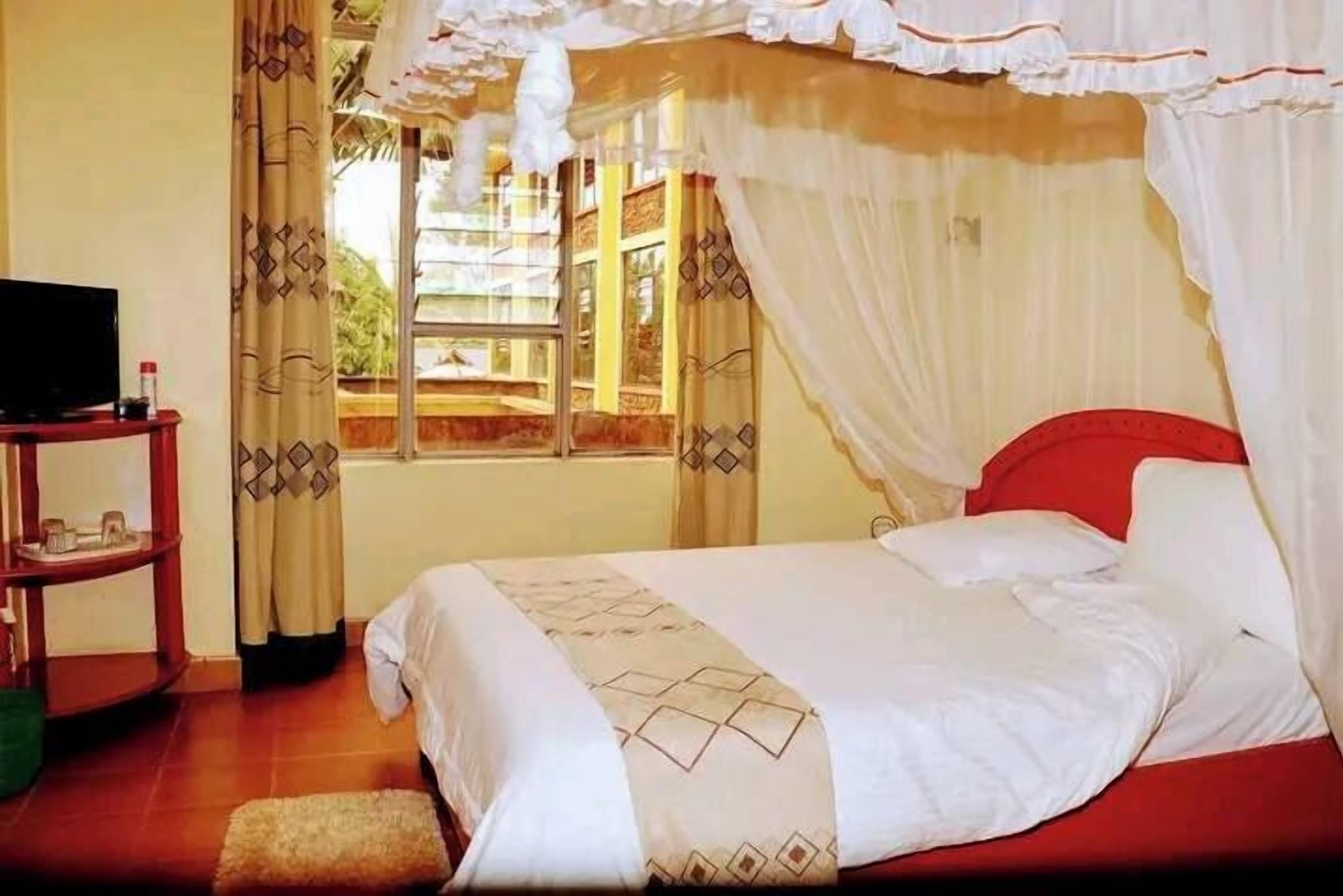 Uma cama com cabeceira vermelha, mesa de cabeceira com televisão e janela com cortinas.