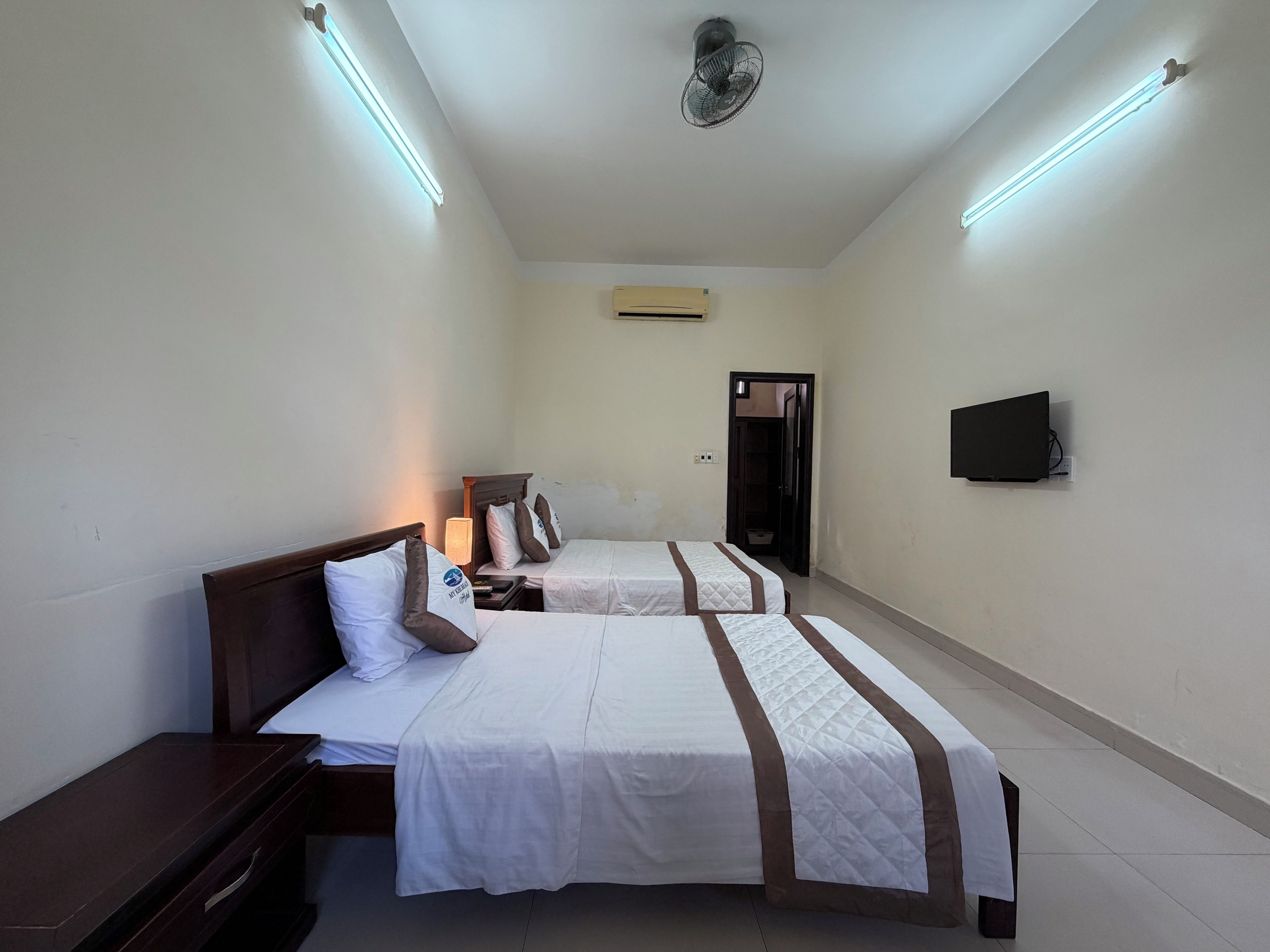 Quarto de hotel com duas camas, uma escrivaninha, luminária, ar condicionado, televisão e ventoinha no teto.
