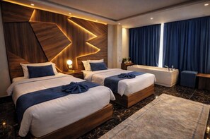 Quarto de hotel com duas camas, área de estar e uma janela grande com cortinas.