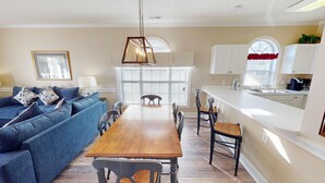 Uma sala de estar moderna com um sofá azul, uma mesa de jantar de madeira e uma área de cozinha com armários brancos e uma janela.