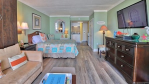Quarto de hotel com cama, sofá, televisão, cômoda e área de cozinha.