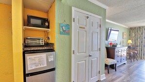 Cozinha compacta com micro-ondas, torradeira e frigorífico, porta fechada com sinalização e área de refeições com mesa e cadeiras.