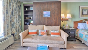 Quarto de hotel com sofá bege, guarda-roupa de madeira, cama com colcha colorida, mesa de cabeceira com abajur e janela com cortinas.