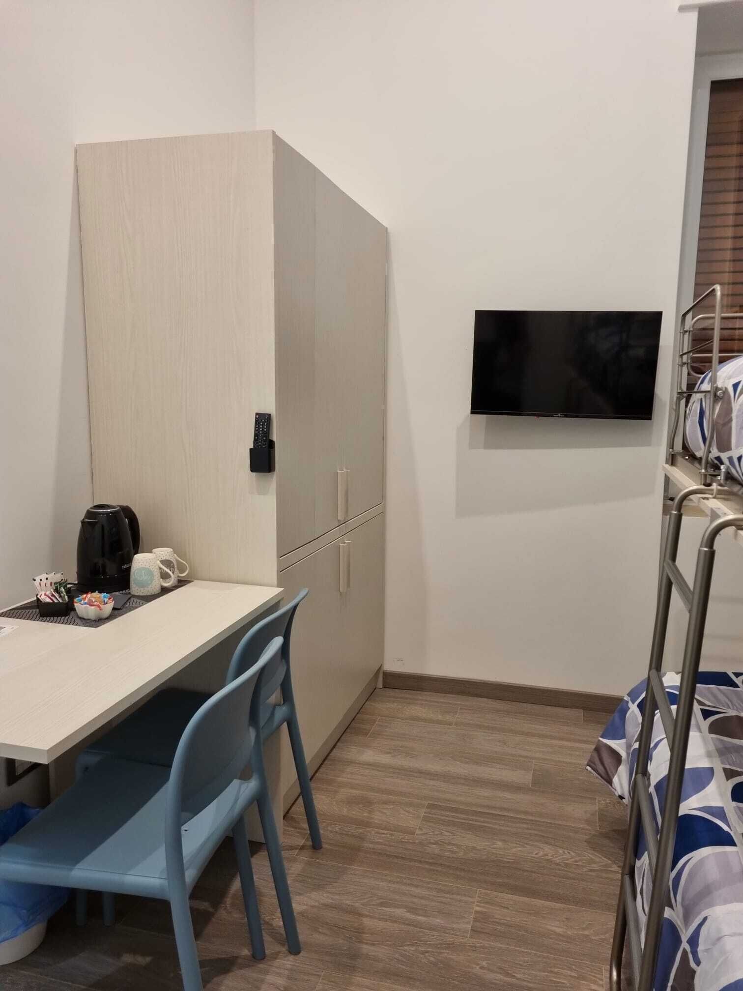 Quarto pequeno e moderno com uma cama, televisão na parede, escrivaninha com cadeira e guarda-roupa.