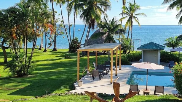 Uma área à beira da piscina com pergolado de madeira, espreguiçadeiras e um pequeno pavilhão com vista para a piscina e o oceano.