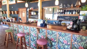 Uma área de bar com banquetas de madeira, um balcão com papel de parede floral tropical e prateleiras cheias de garrafas e copos.
