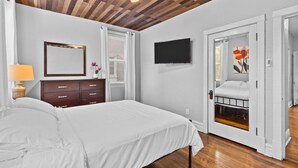 Quarto com cama, teto de madeira, televisão, penteadeira e porta para outro cômodo.