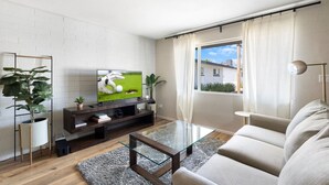 Uma sala de estar moderna com uma TV de tela plana, uma mesa de centro de vidro, um sofá bege e vista para uma área residencial pela janela.