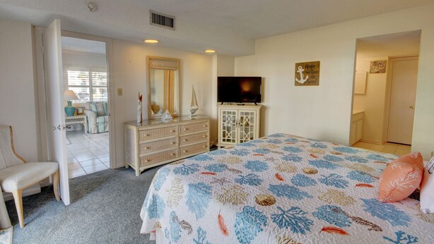 Quarto com cama, penteadeira, televisão e porta que dá para outro cômodo.