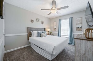 Um quarto com cama, ventilador de teto, mesa de cabeceira, luminária e cômoda.