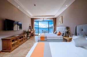 Quarto de hotel com uma cama grande, televisão, armário de madeira, escrivaninha com cadeira e vista para o mar através de uma janela ampla.