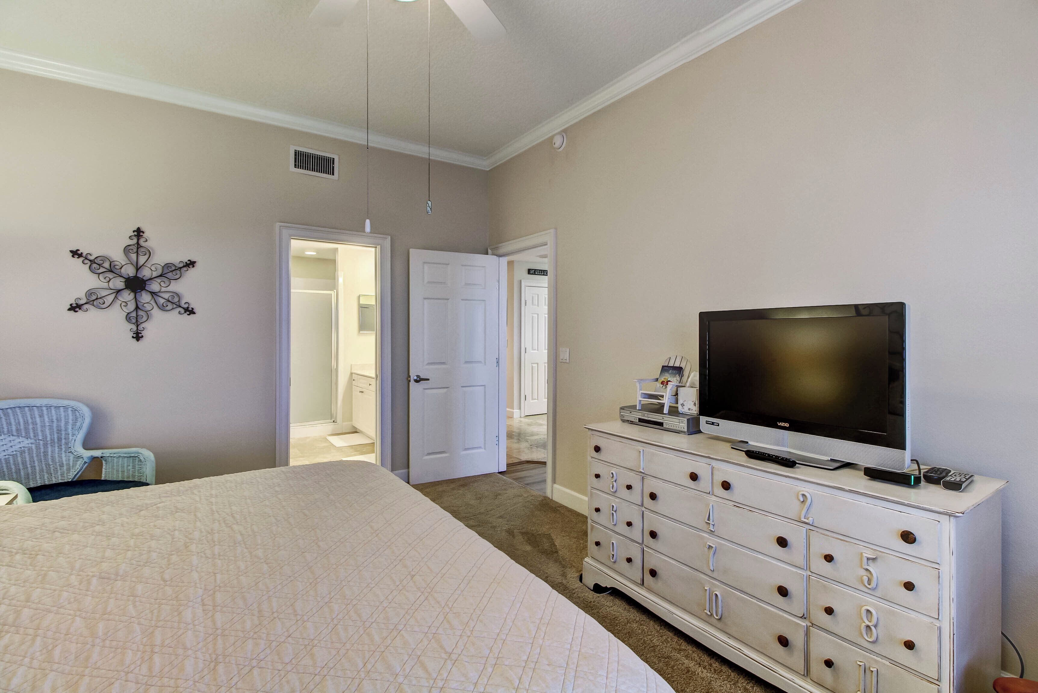 Quarto com cama, penteadeira, televisão e um objeto decorativo na parede.