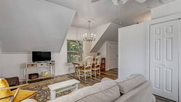 Uma sala de estar espaçosa com um sofá branco, uma mesa de jantar com cadeiras, um lustre e uma televisão sobre um móvel.