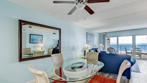 Uma sala de estar com uma mesa de vidro, cadeiras de vime, um sofá azul e vista para o oceano.