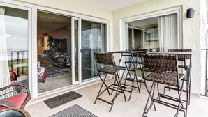 Uma varanda com mesa e cadeiras, uma porta de correr de vidro que dá para um espaço interior e um tapete pequeno no chão.