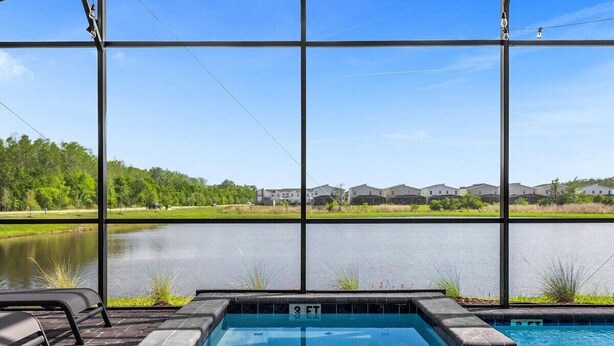 Piscina com hidromassagem, rodeada por um terraço de tijolos e grandes janelas com vista para um lago e edifícios distantes.