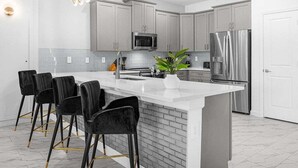 Uma cozinha moderna com ilha central, banquetas, eletrodomésticos de aço inoxidável e painel de fundo branco.