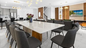 Uma área de refeições moderna, com uma grande mesa retangular, cadeiras estofadas em cinza e iluminação pendente.