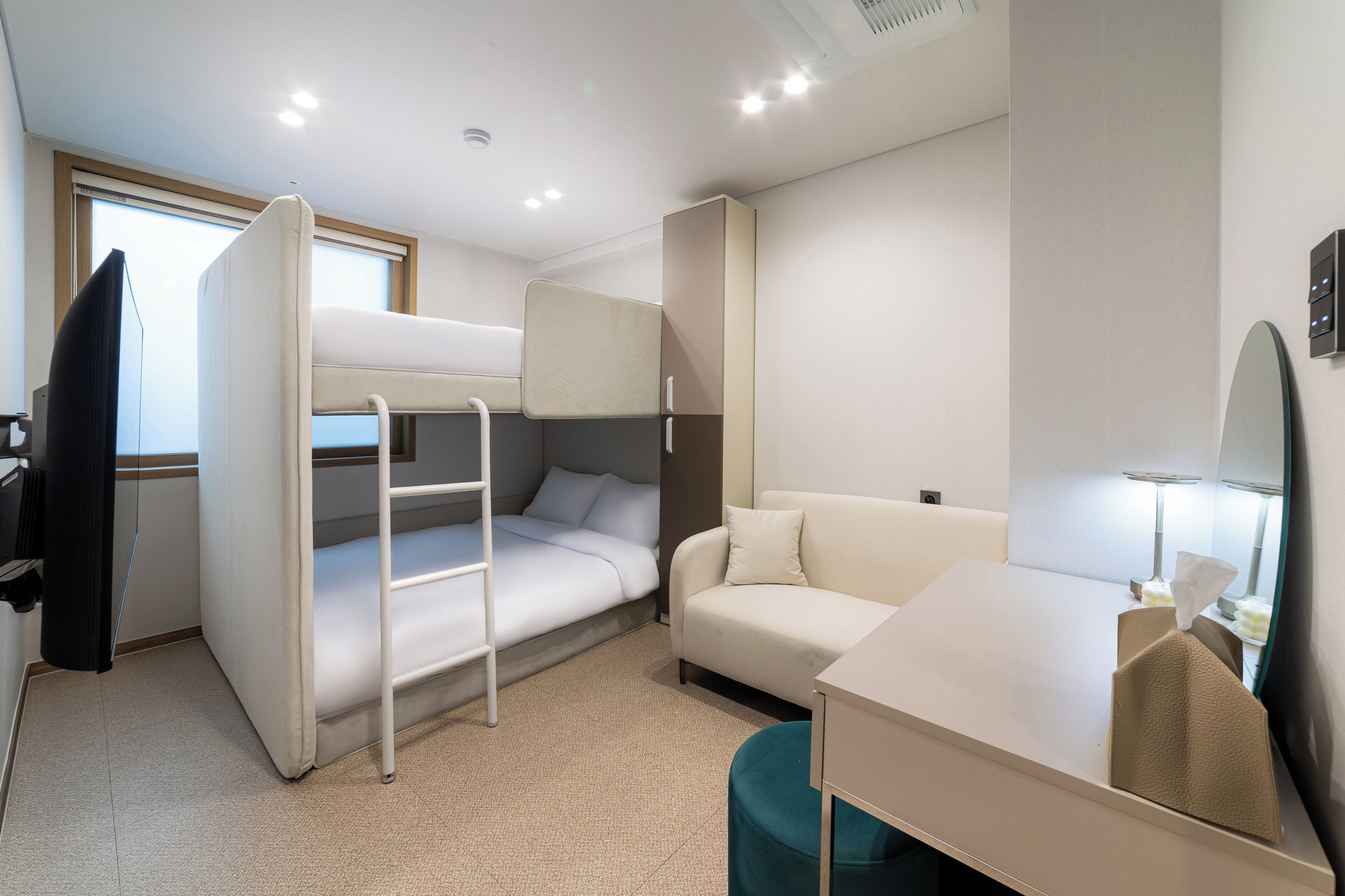 Quarto de hotel compacto com beliche, sofá, escrivaninha e espelho.