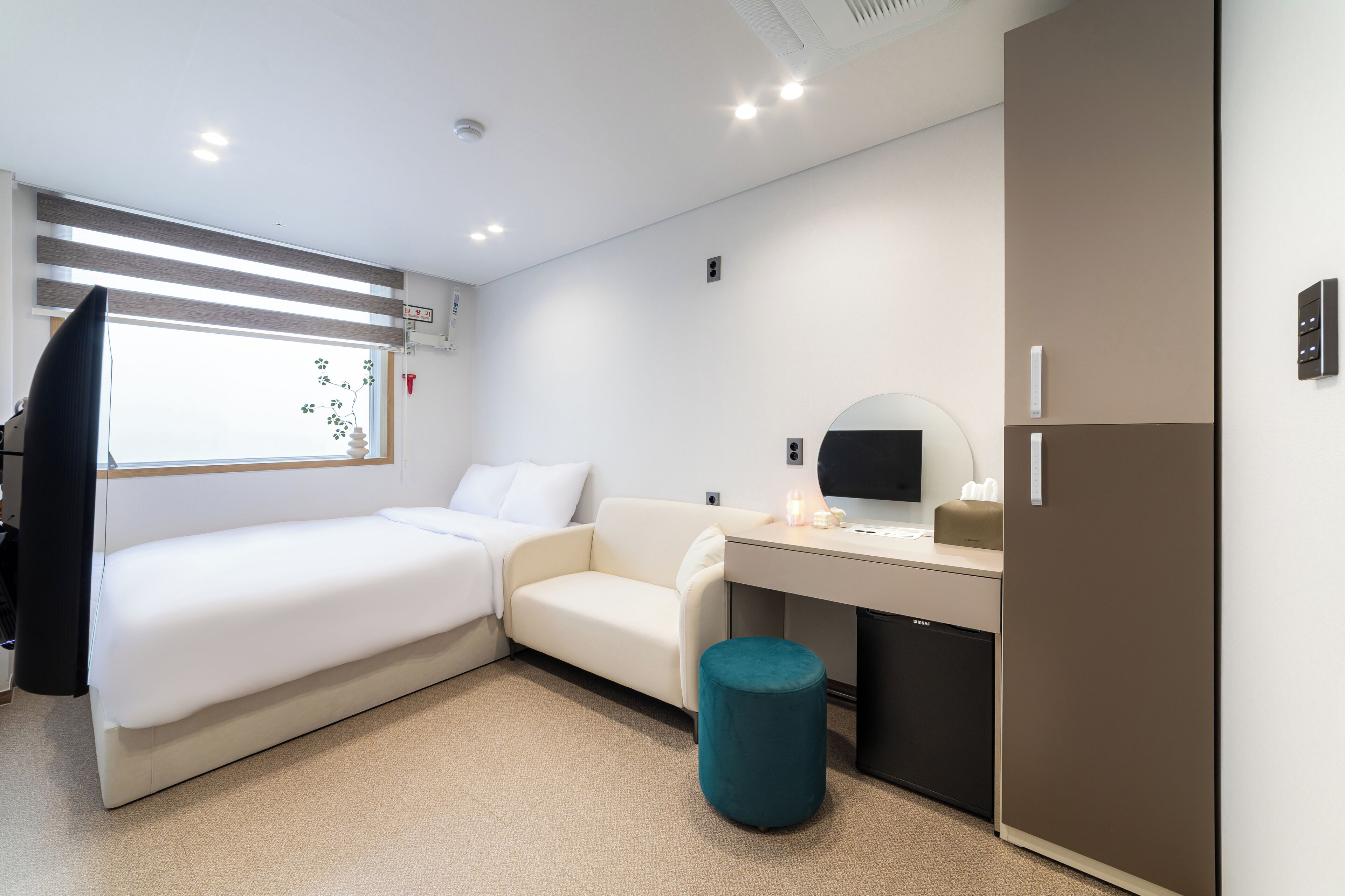 Quarto de hotel moderno com cama, escrivaninha, cadeira, um banquinho pequeno, televisão e guarda-roupa.