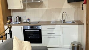 Uma cozinha compacta com armários brancos, coifa de aço inoxidável, forno embutido, pia e uma pequena lixeira.