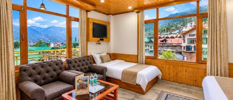 Quarto de hotel com cama, sofá, televisão e vista para montanhas e uma aldeia.