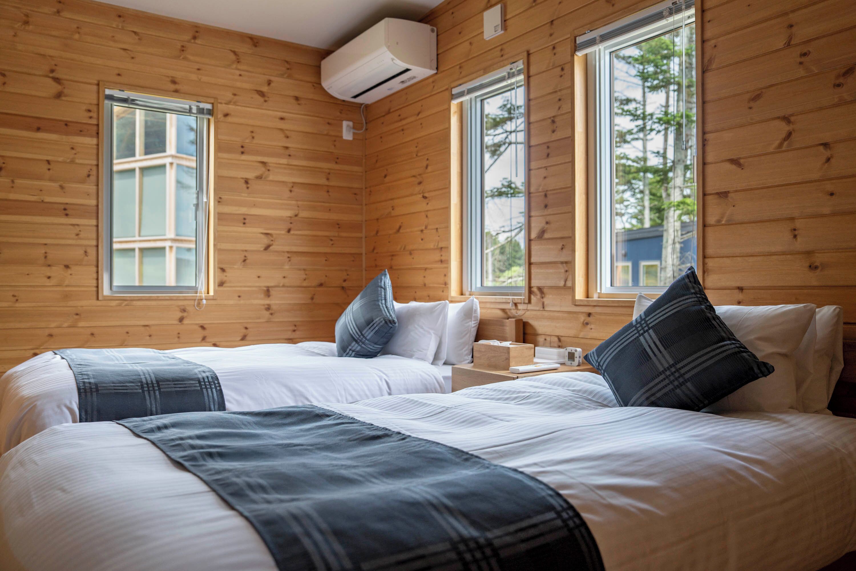 Quarto com paredes de madeira, duas camas com roupa de cama branca e azul, e grandes janelas com vista para árvores.