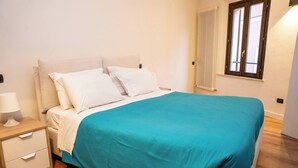 Quarto com cama, mesa de cabeceira, abajur e janela com persianas.