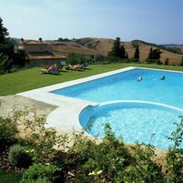 Uma piscina retangular com uma seção curva, rodeada por um deck de pedra. Há espreguiçadeiras e um pequeno edifício com telhado de azulejos ao fundo.
