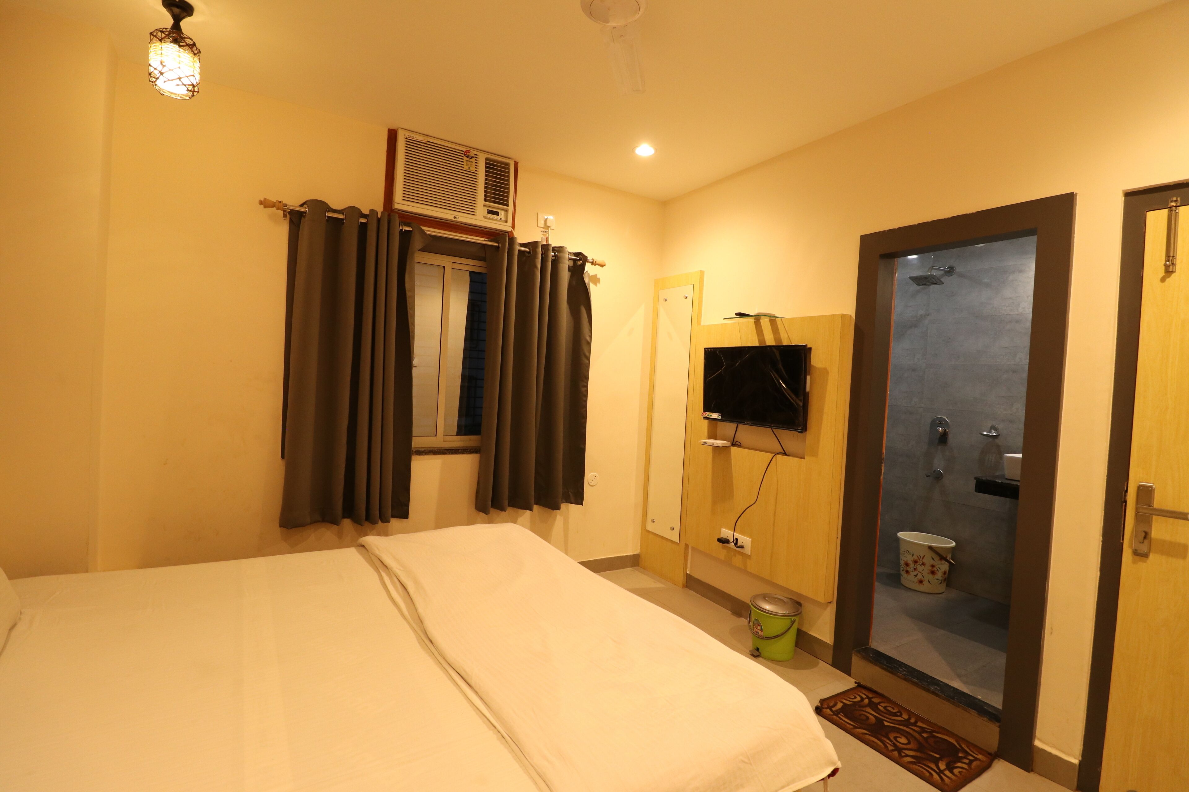 Quarto de hotel com cama, televisão na parede, área de chuveiro e uma pequena janela com cortinas.