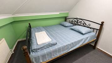 Uma cama de solteiro com estrutura de metal e colcha listrada em azul, uma toalha branca e duas almofadas num quarto com paredes verdes e teto inclinado.