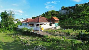 Uma casa de dois andares com telhado vermelho, rodeada por vegetação exuberante e um pequeno jardim.
