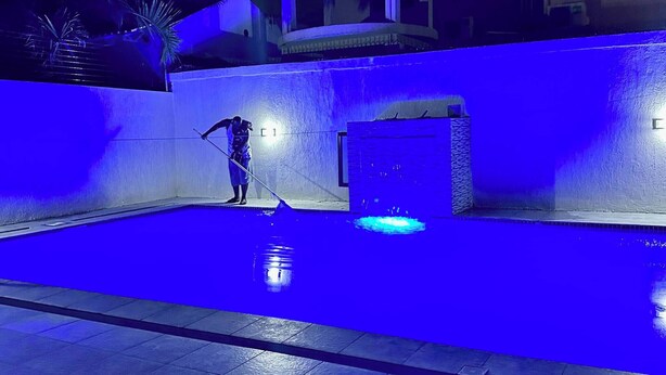 Vista noturna de uma piscina iluminada em azul, uma pessoa com guarda-chuva e um edifício ao fundo.