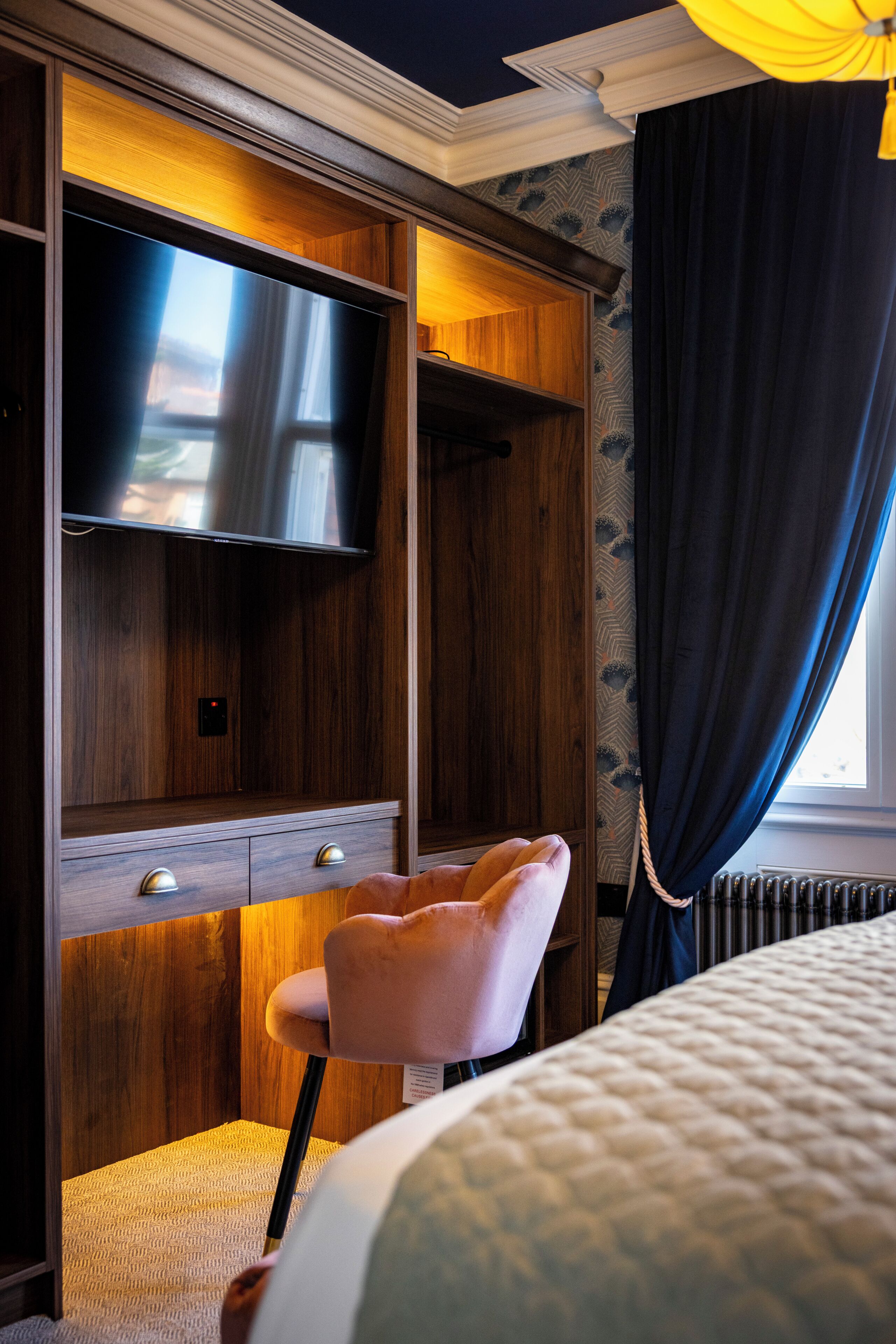 Quarto com cama, televisão presa em parede de madeira, uma cadeira e janela com cortinas.