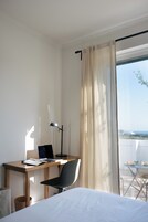 Quarto de hotel com cama, escrivaninha com laptop, cadeira e vista para o exterior através de uma grande janela.