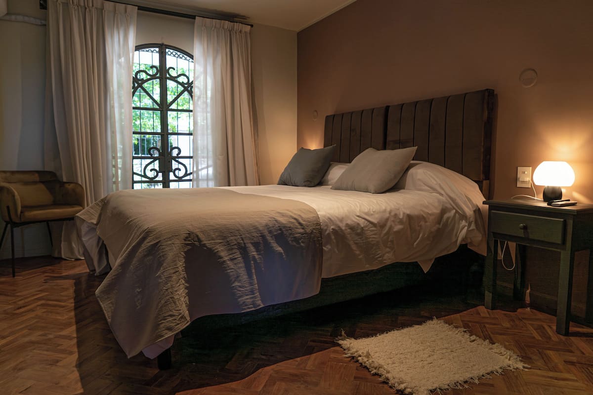 Uma cama bem arrumada com cabeceira de madeira, mesa de cabeceira com abajur e uma janela com cortinas.